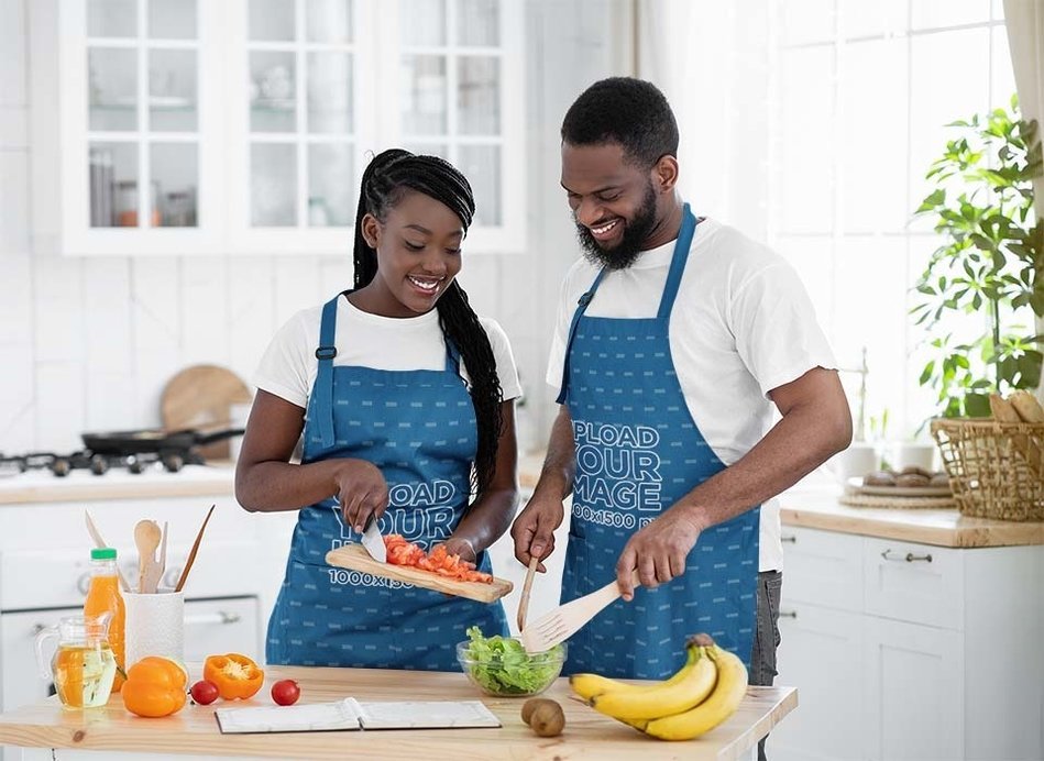 Placeit - Mockup of a Smiling Couple Cooking in Matching Sublimated Aprons