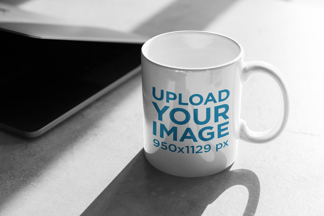 Placeit - 11 oz Coffe Mug Mockup Beside a Semi-Open Laptop
